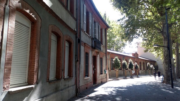 Rua com edifícios com janelas em arco e venezianas, calçada e árvores que oferecem sombra.