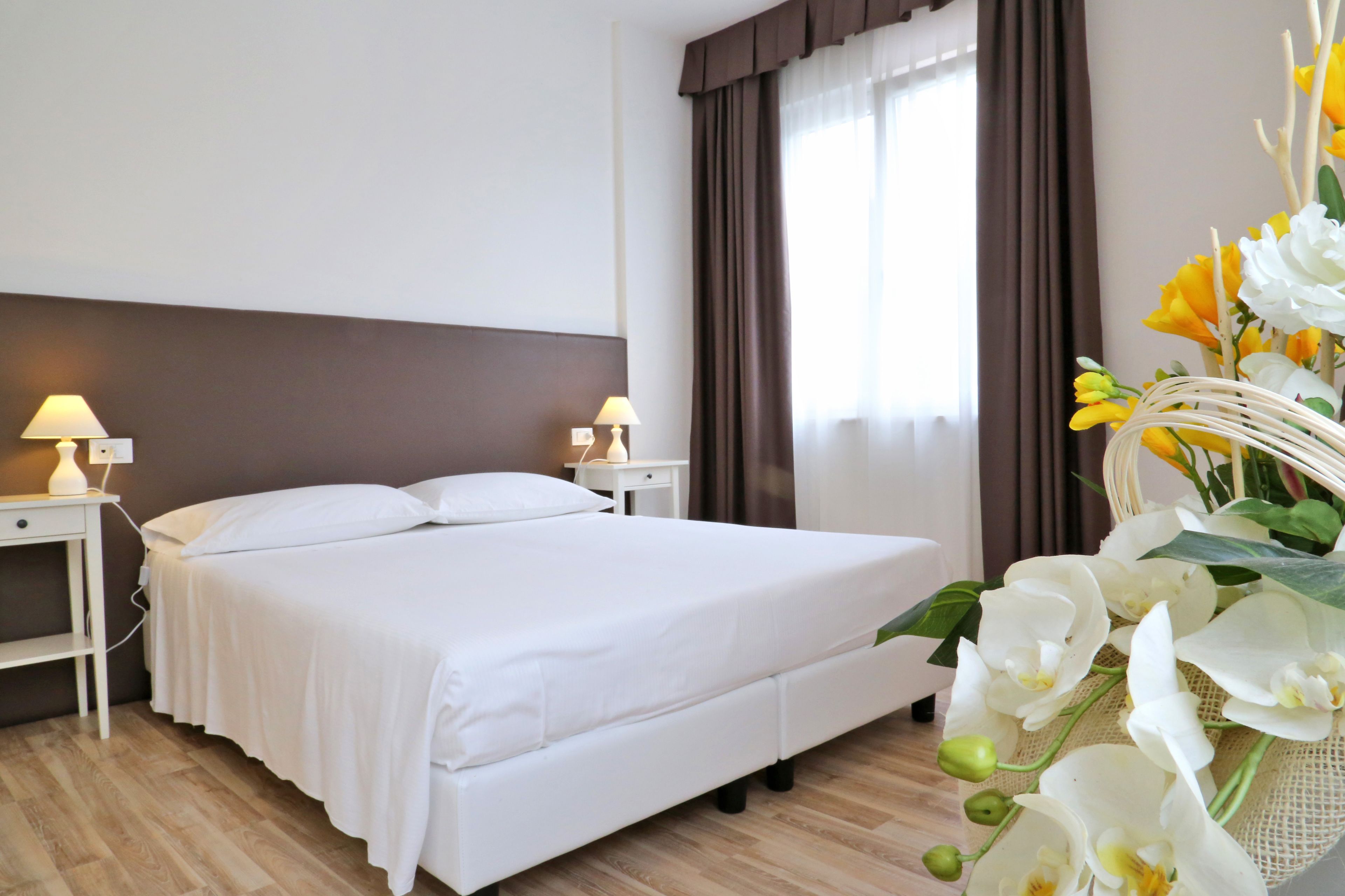 Quarto de hotel com uma cama grande, duas mesas de cabeceira com abajures, uma janela com cortinas e um arranjo floral.