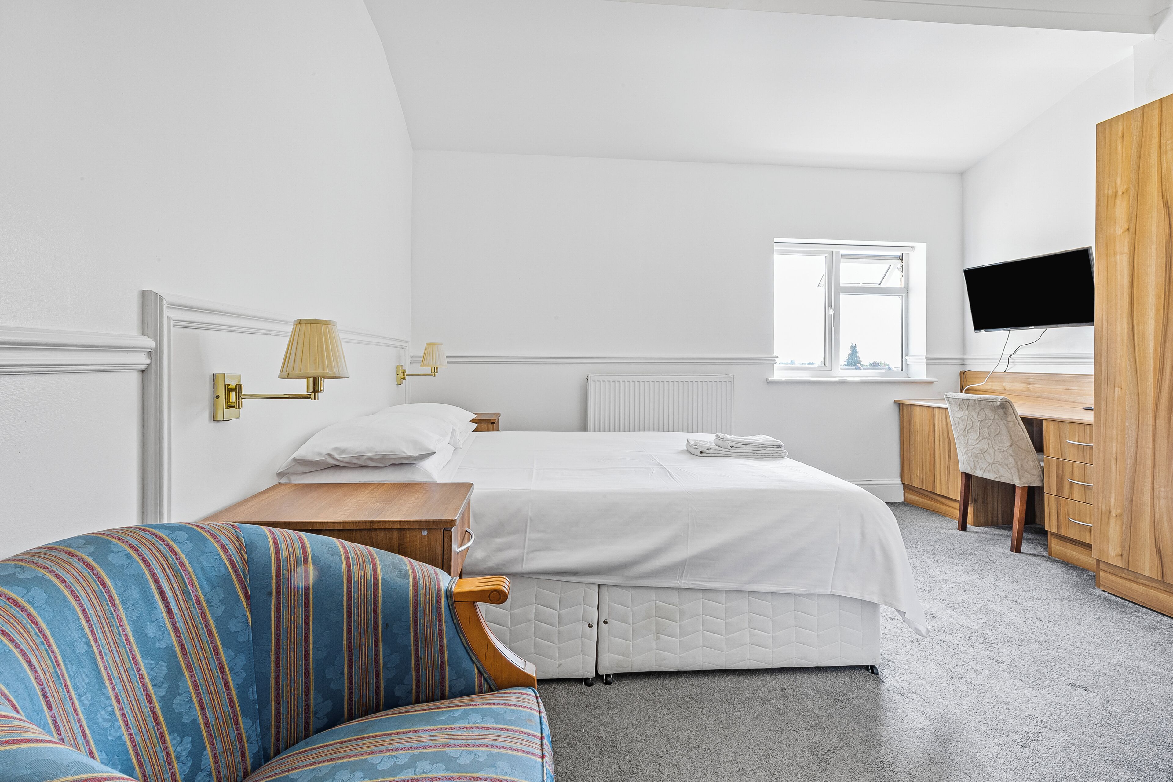 Quarto de hotel com cama, escrivaninha, cadeira, televisão e janela.