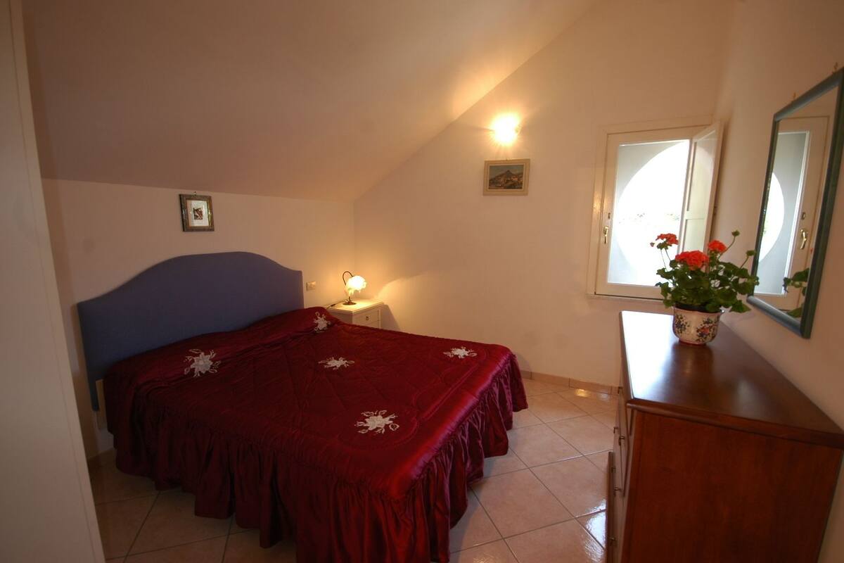 Quarto com cama, mesa de cabeceira, abajur e um vaso de planta.