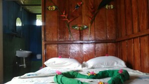 Uma cama de madeira com almofadas brancas e um cobertor verde, uma cabeceira de madeira com design floral, e uma pia no quarto vizinho.