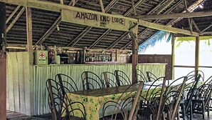 Uma área de refeições rústica ao ar livre, com mesa e cadeiras, uma placa indicando "Amazon King Lodge" e um bar com garrafas e sinalização de serviço 24 horas.