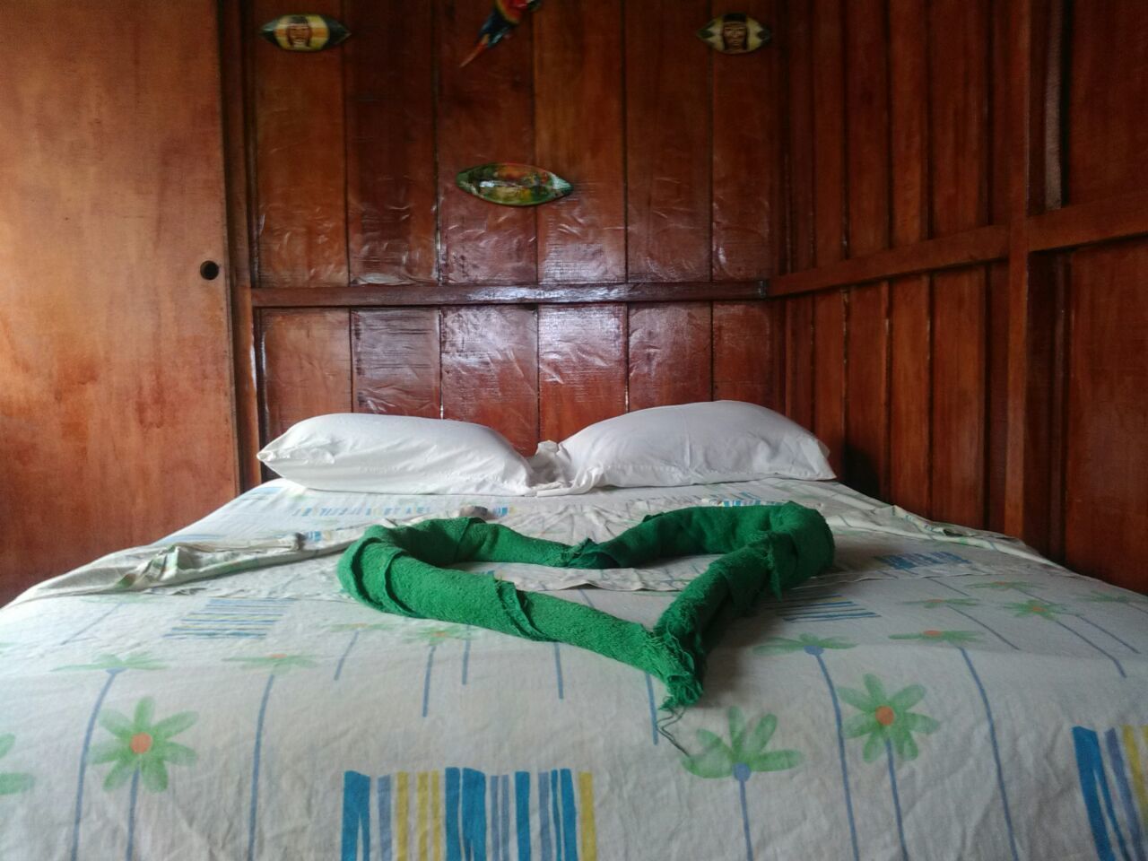 Uma cama com um cobre-leito estampado com flores, duas almofadas brancas e um objeto em formato de coração verde sobre a cama.
