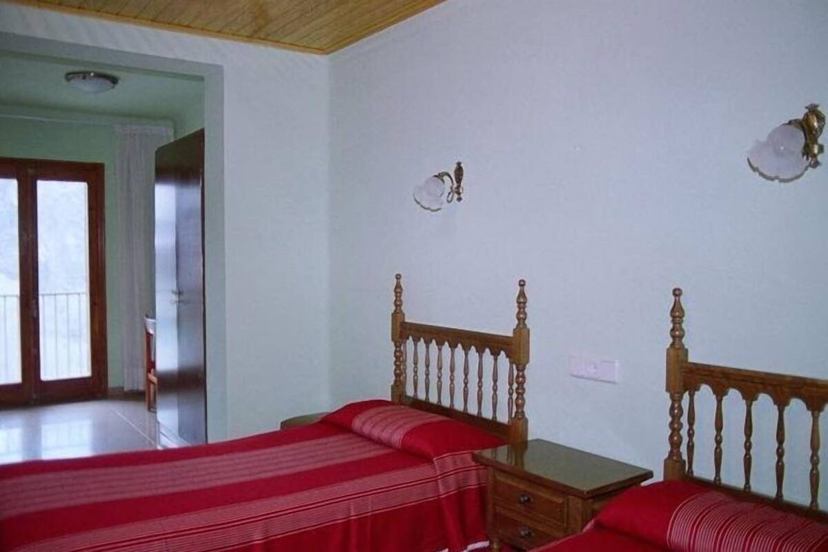 Quarto com duas camas de madeira, uma mesinha de cabeceira e uma janela com vista.