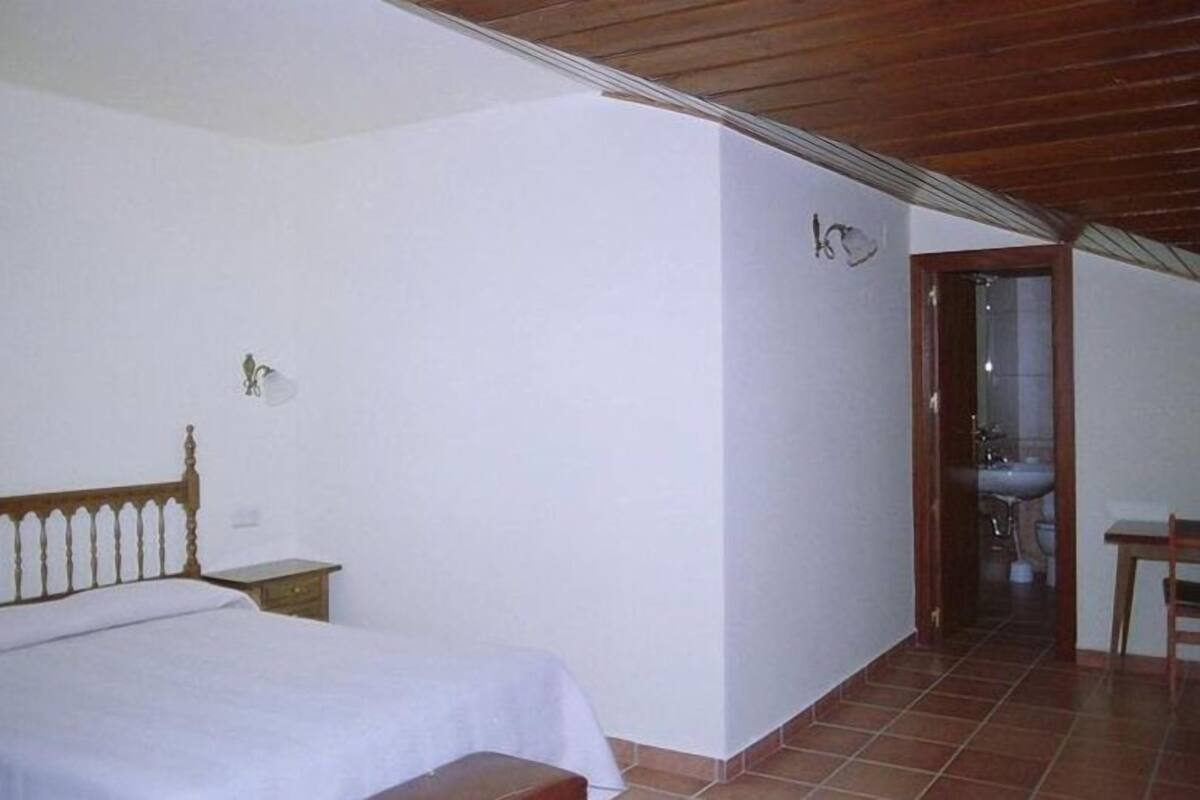 Quarto com cama, mesa de cabeceira, porta de madeira e banheiro visível através de uma porta aberta.