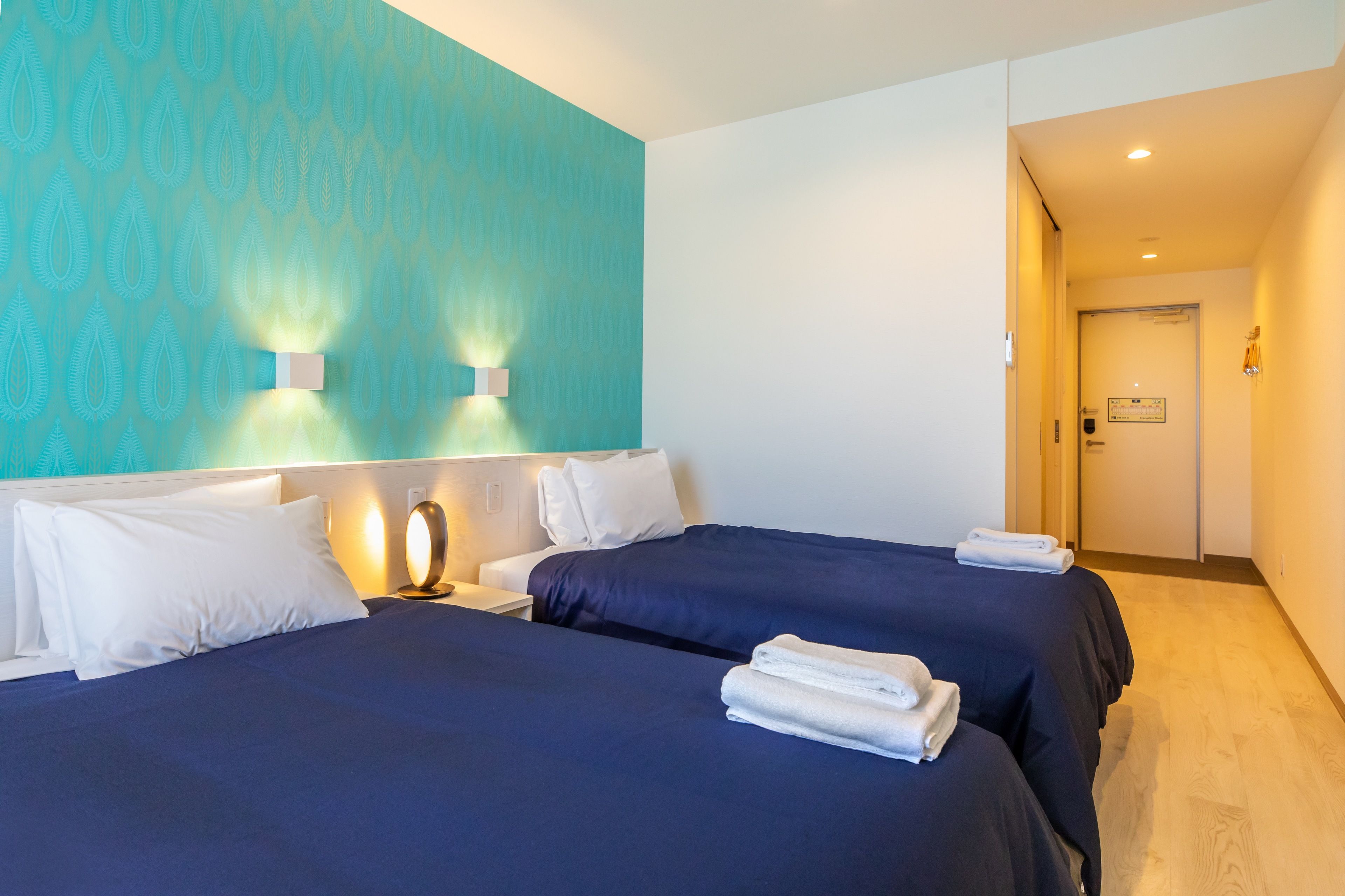 Quarto de hotel com duas camas, papel de parede estampado em tons de verde-água e uma porta que dá para um corredor.