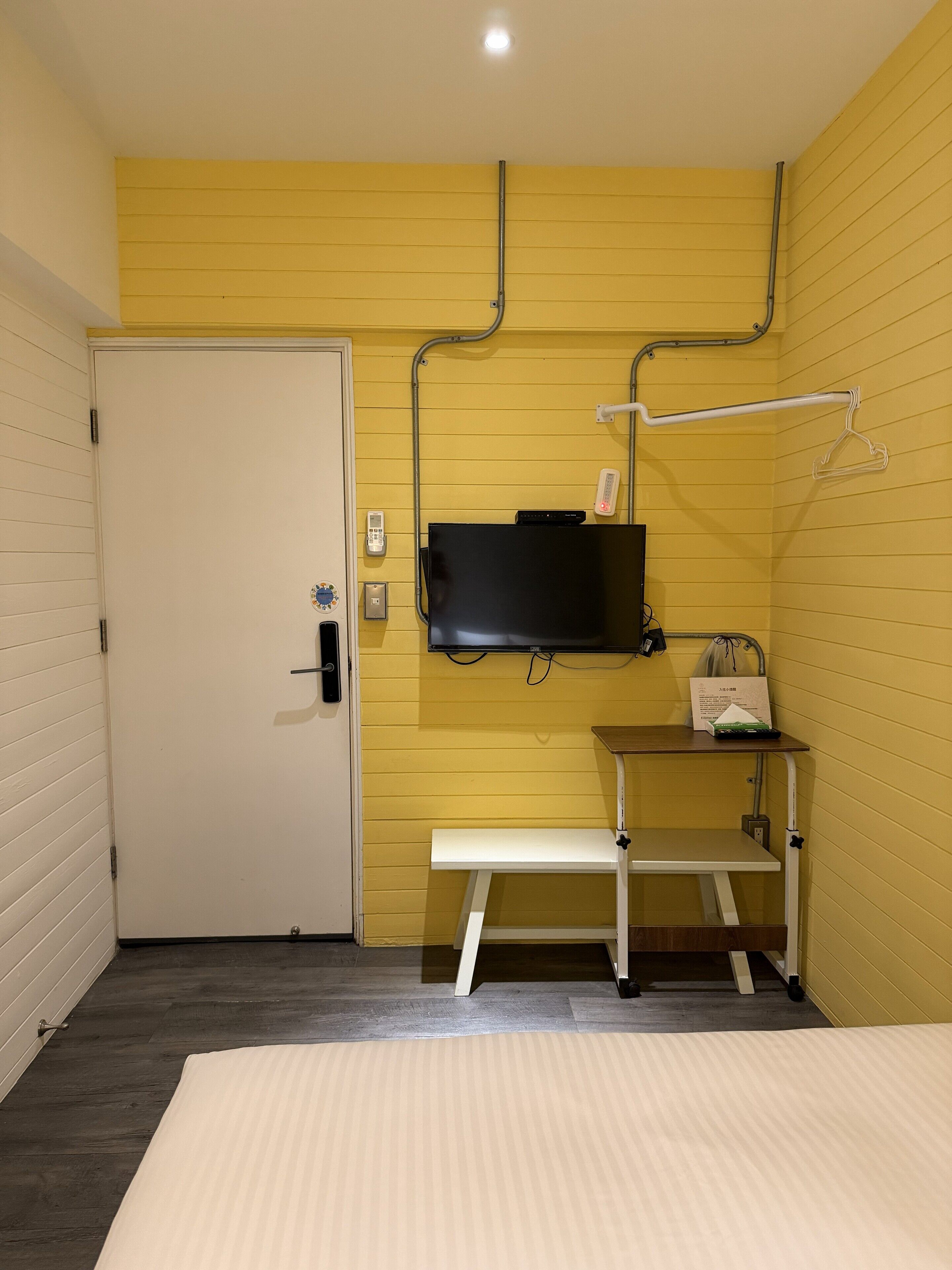 Quarto de hotel com cama, uma mesinha, televisão na parede e uma porta.