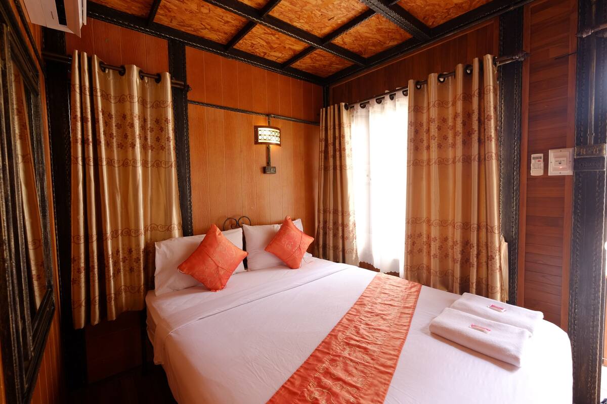 Uma cama bem arrumada com roupa de cama branca e laranja, uma luminária fixada na parede e cortinas em paredes de madeira.