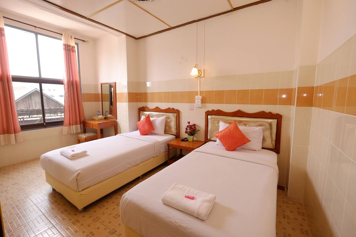 Quarto de hotel com duas camas, uma janela com cortinas, uma mesa de cabeceira com abajur e vista para um edifício.