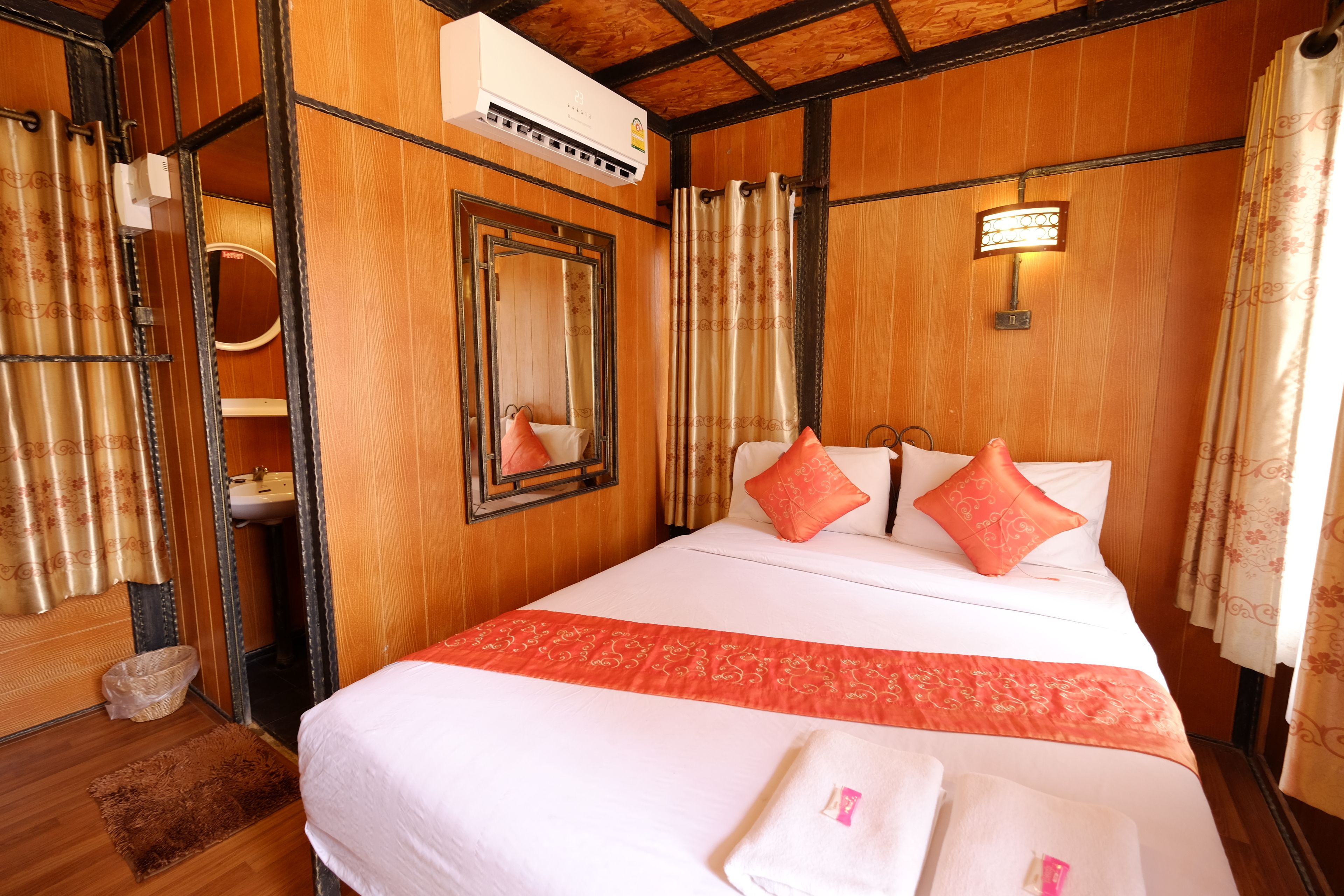 Quarto de hotel com decoração em madeira, uma cama com roupa de cama branca e almofadas laranja, um espelho, uma pia e um aplique de parede.