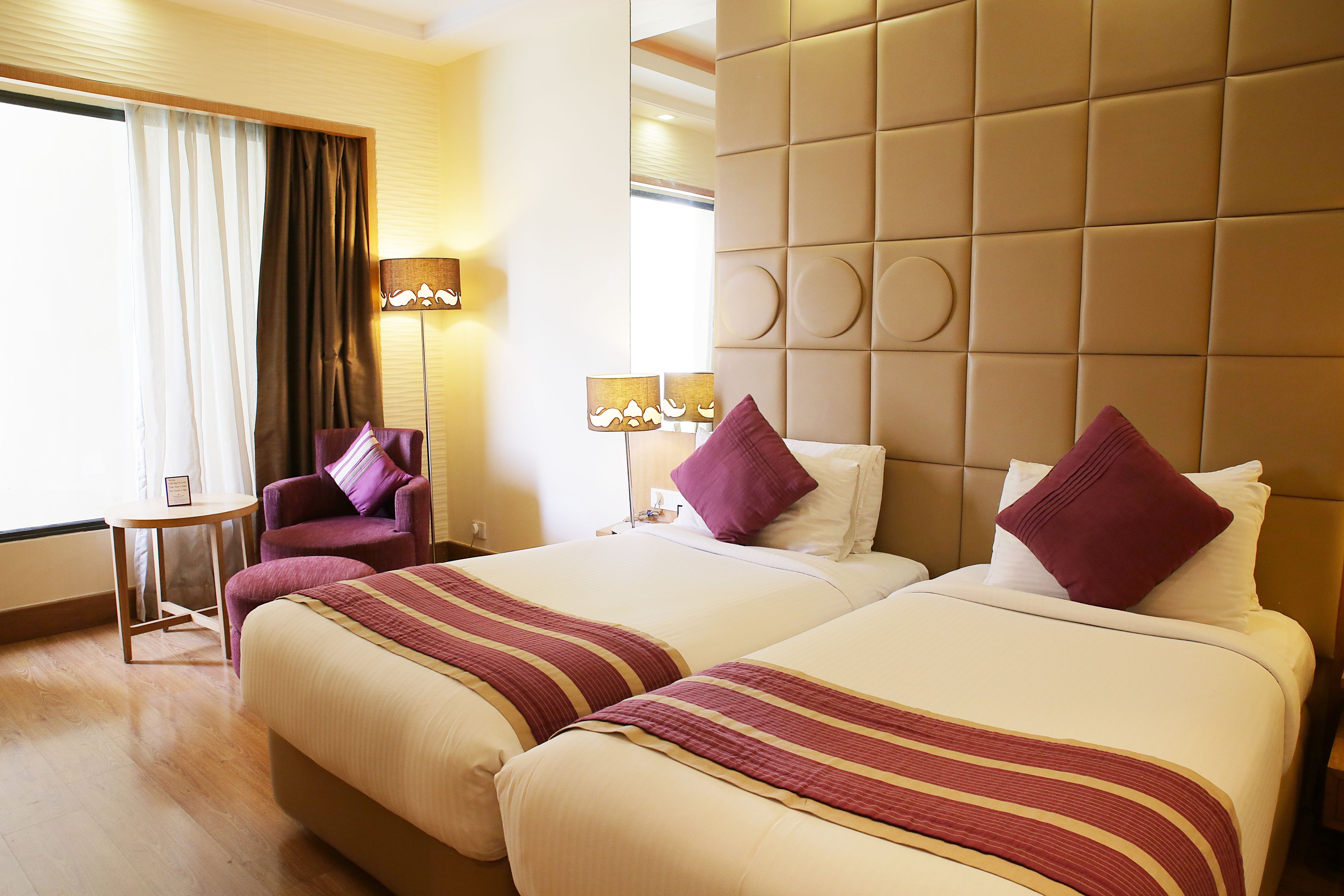 Quarto de hotel com duas camas, uma cadeira, uma mesinha e um abajur.