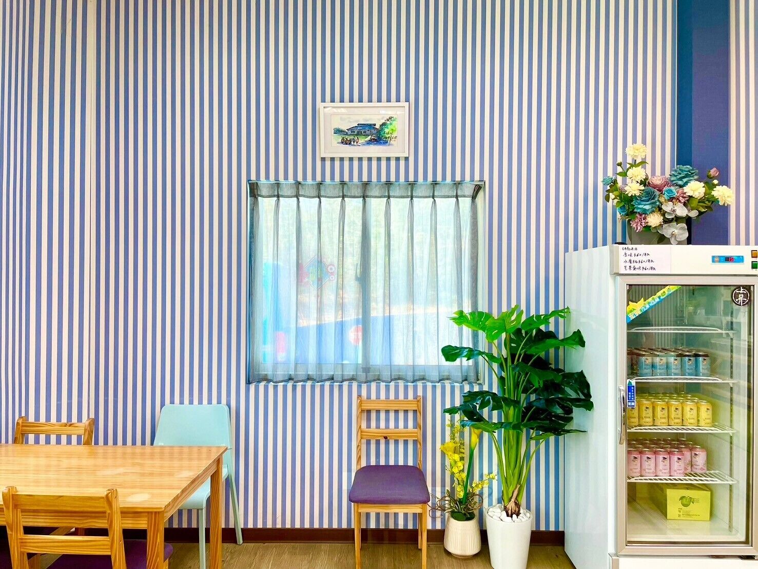 Um quarto com papel de parede listrado, uma janela com cortinas finas, uma mesa de madeira com cadeiras, um refrigerador cheio de bebidas e duas plantas em vasos.