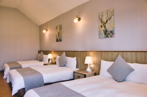 Quarto de hotel com três camas, cada uma com uma mesa de cabeceira e abajur. Há luminárias fixadas na parede e duas pinturas na parede.
