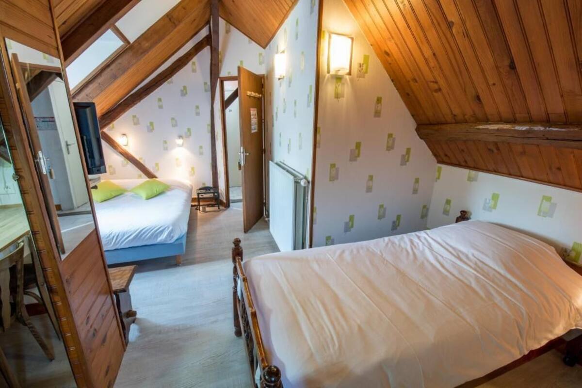 Quarto com teto inclinado, vigas de madeira, cama com colcha branca, uma mesinha e uma cadeira.