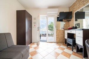 Quarto de hotel com sofá, escrivaninha com cadeira, televisão na parede e porta de vidro que dá para área externa com mesa e cadeiras.