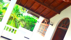 Área externa coberta com ventilador de teto, porta de madeira e vista para palmeiras.