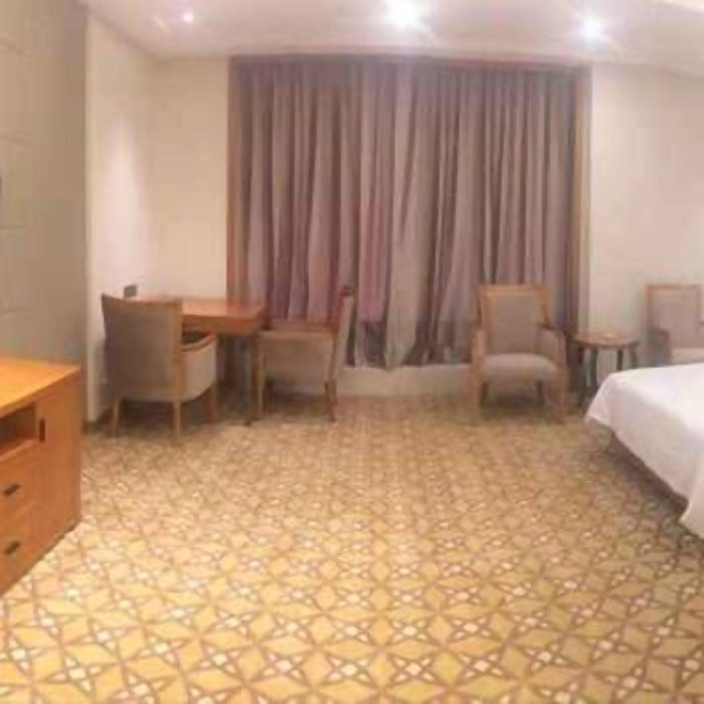 Quarto de hotel com cama, escrivaninha, duas cadeiras, uma mesinha e chão estampado.
