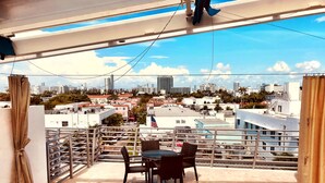 Uma varanda com mesa e cadeiras, vista para um horizonte urbano com edifícios e uma bandeira.