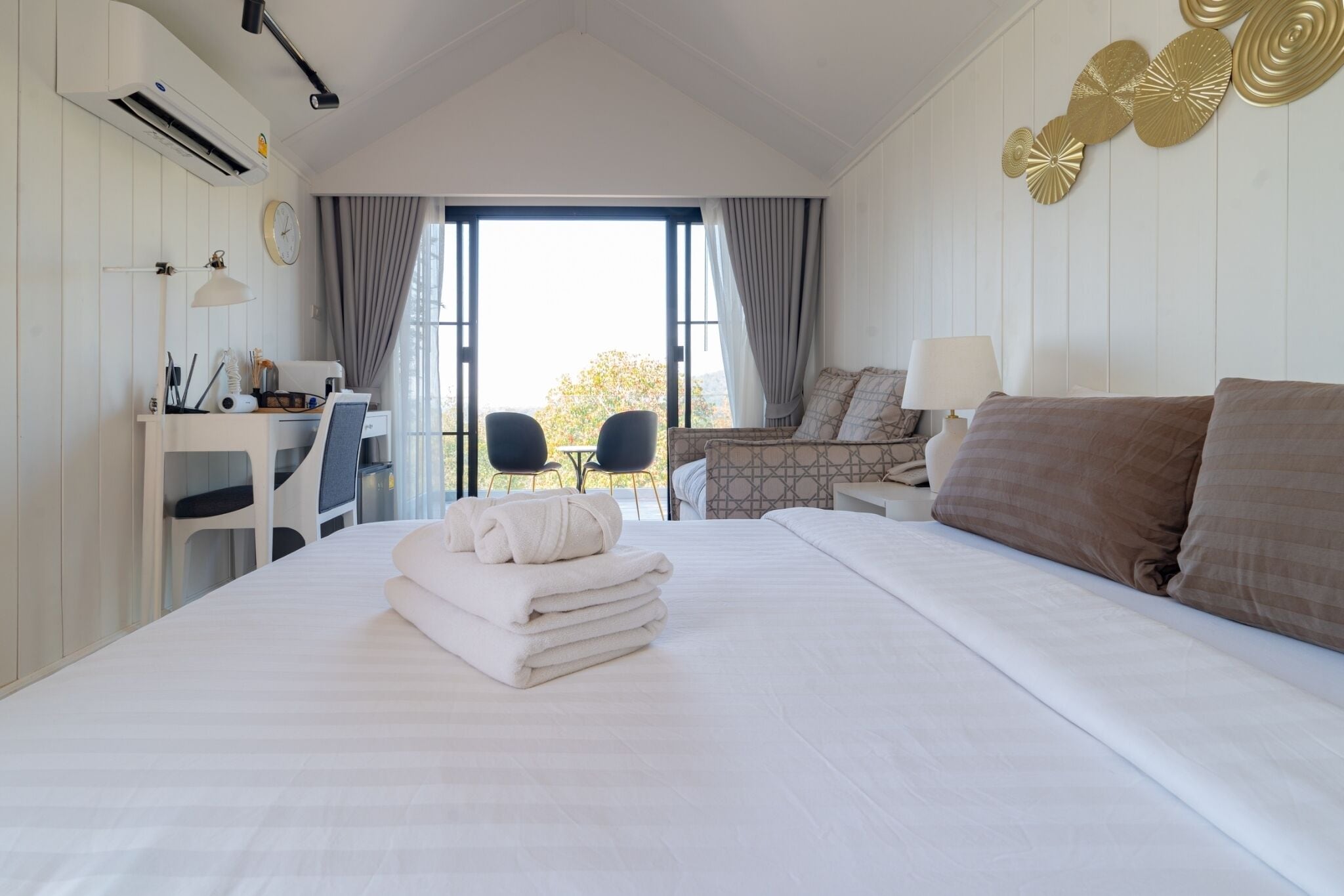 Quarto de hotel com cama, escrivaninha, cadeira, mesa pequena e janela com cortinas.