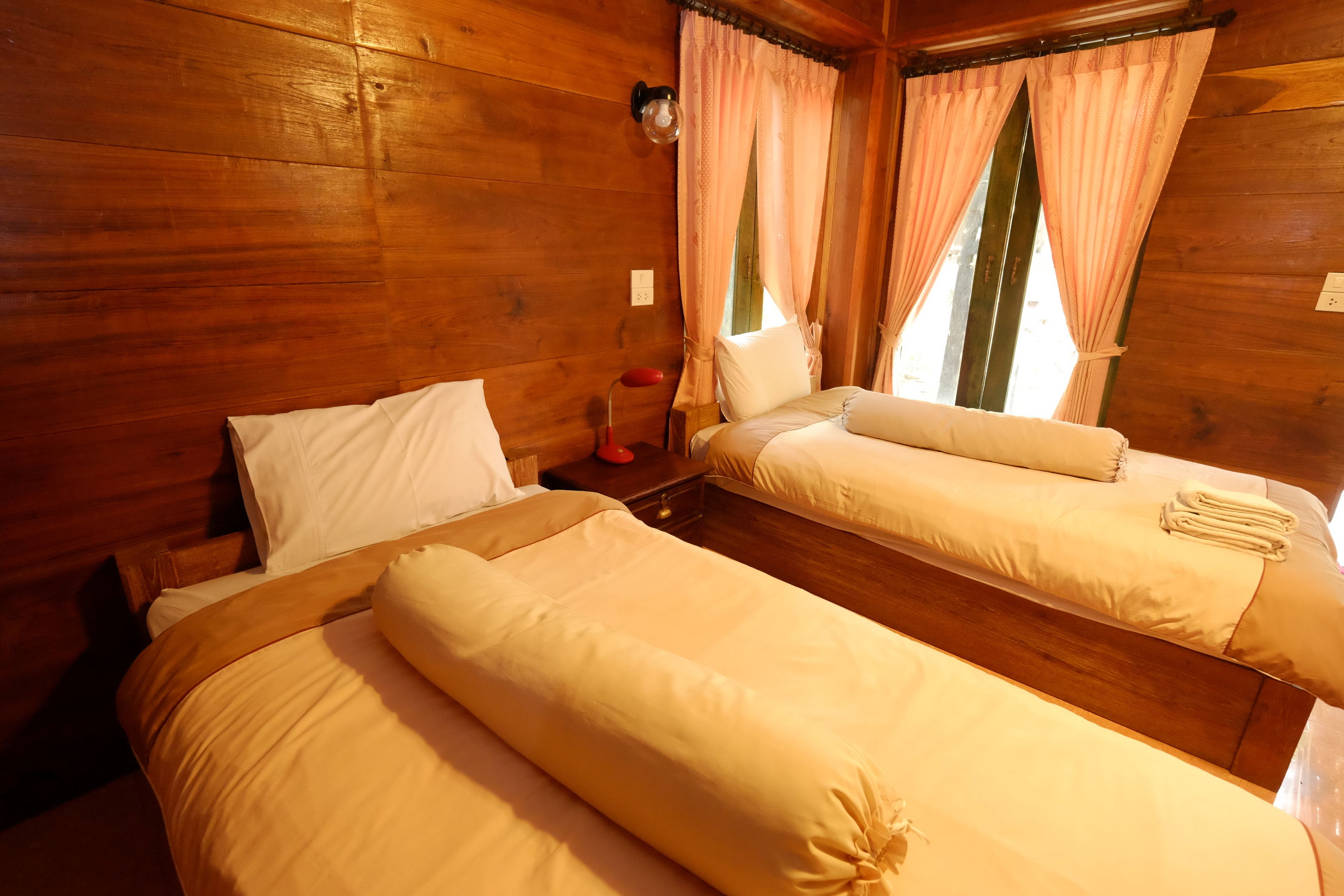 Quarto de cabana de madeira com duas camas, cada uma com roupa de cama e almofadas brancas. Há uma mesa de cabeceira com abajur e uma pequena mesa com um objeto vermelho.