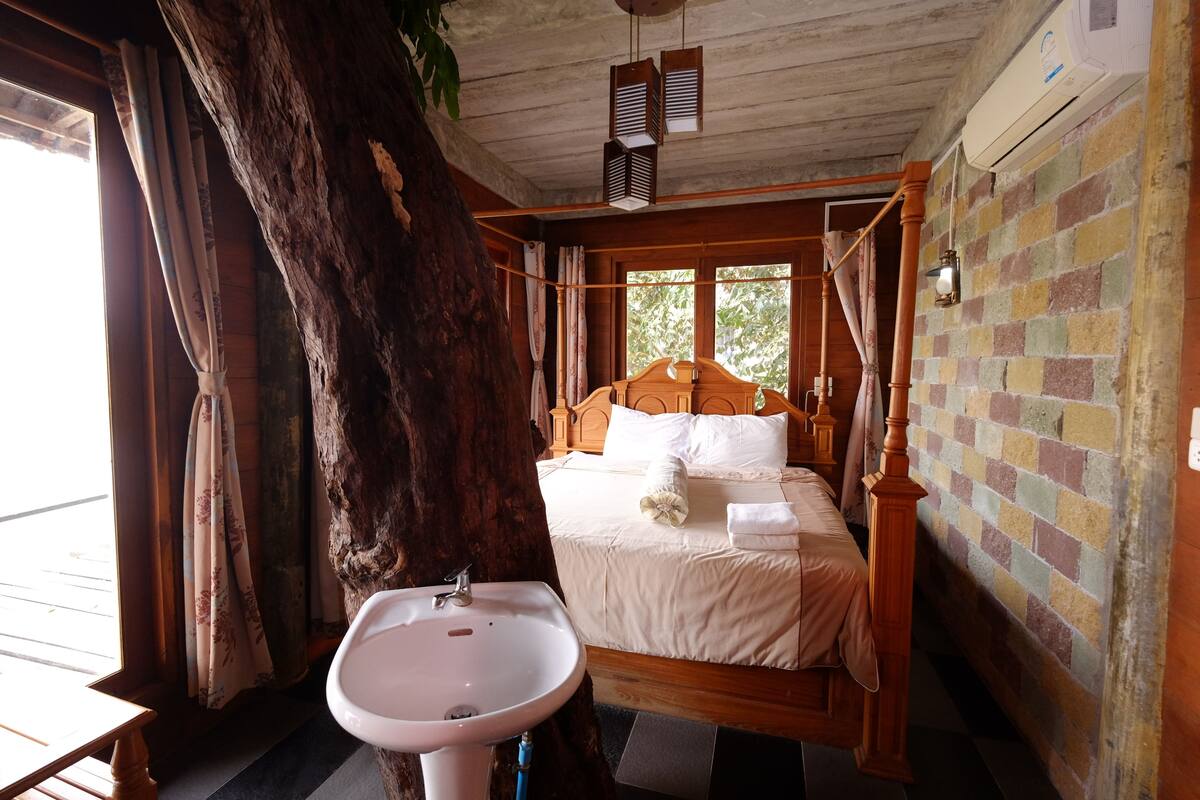 Quarto com cama de madeira, pia branca e um grande tronco de árvore incorporado ao design do espaço.