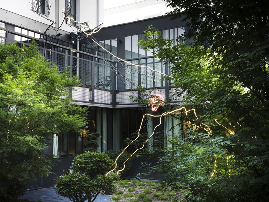 Um edifício moderno com fachada de vidro, rodeado por vegetação exuberante e uma instalação de luz decorativa.