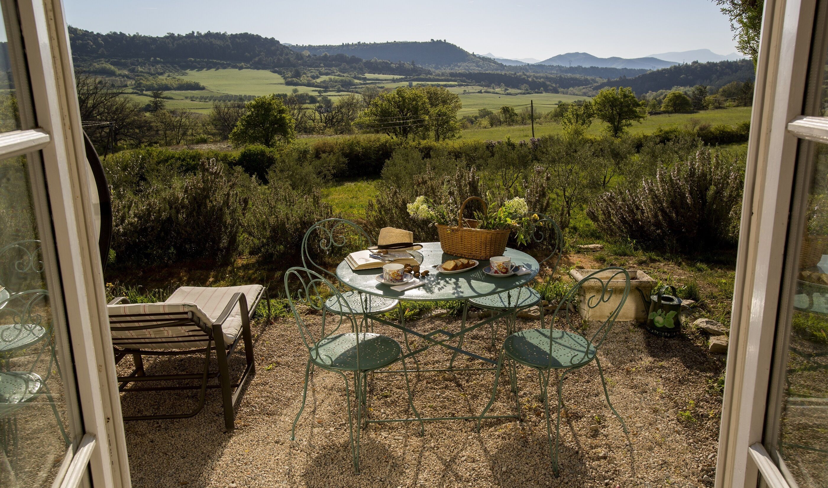 Um pátio com mesa posta para refeição, rodeado de cadeiras, com vista para uma paisagem campestre.