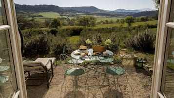 Um pátio com mesa posta para refeição, rodeado de cadeiras, com vista para uma paisagem campestre.