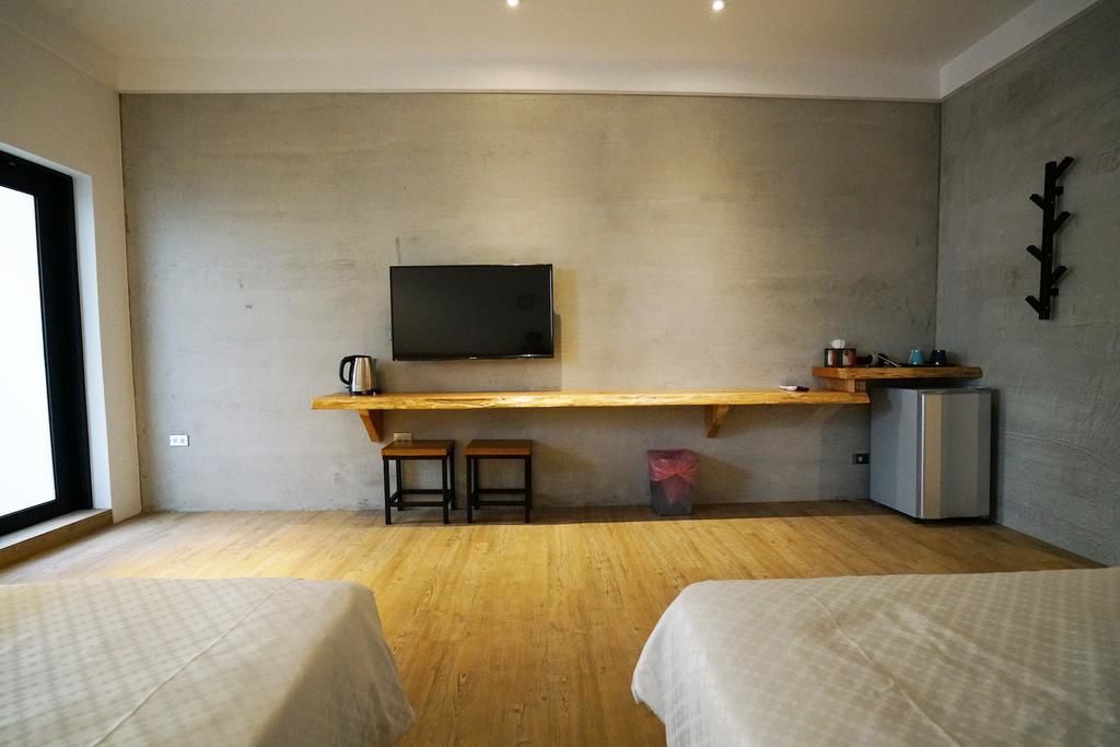 Quarto com piso de madeira, duas camas, televisão de tela plana na parede, uma pequena cozinha compacta com geladeira e uma janela que proporciona iluminação natural.