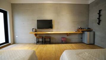 Quarto com piso de madeira, duas camas, televisão de tela plana na parede, uma pequena cozinha compacta com geladeira e uma janela que proporciona iluminação natural.