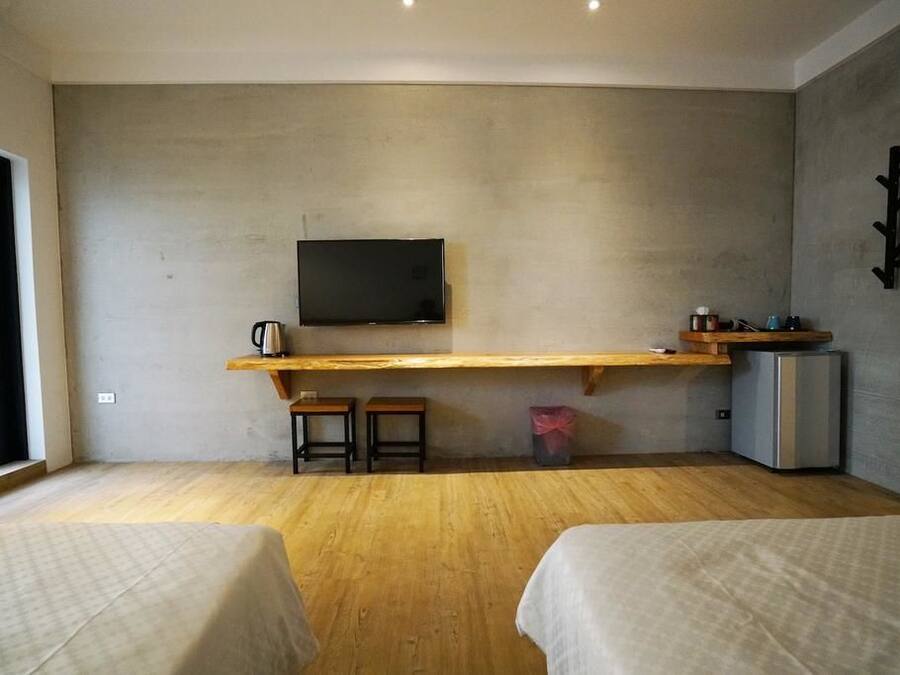 Quarto com piso de madeira, duas camas, televisão de tela plana na parede, uma pequena cozinha compacta com geladeira e uma janela que proporciona iluminação natural.
