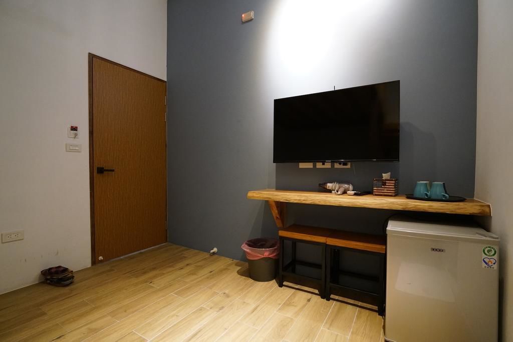 Um quarto pequeno com piso de madeira, um banco de madeira, uma televisão de tela plana fixada na parede e uma geladeira pequena.