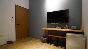 Um quarto pequeno com piso de madeira, um banco de madeira, uma televisão de tela plana fixada na parede e uma geladeira pequena.