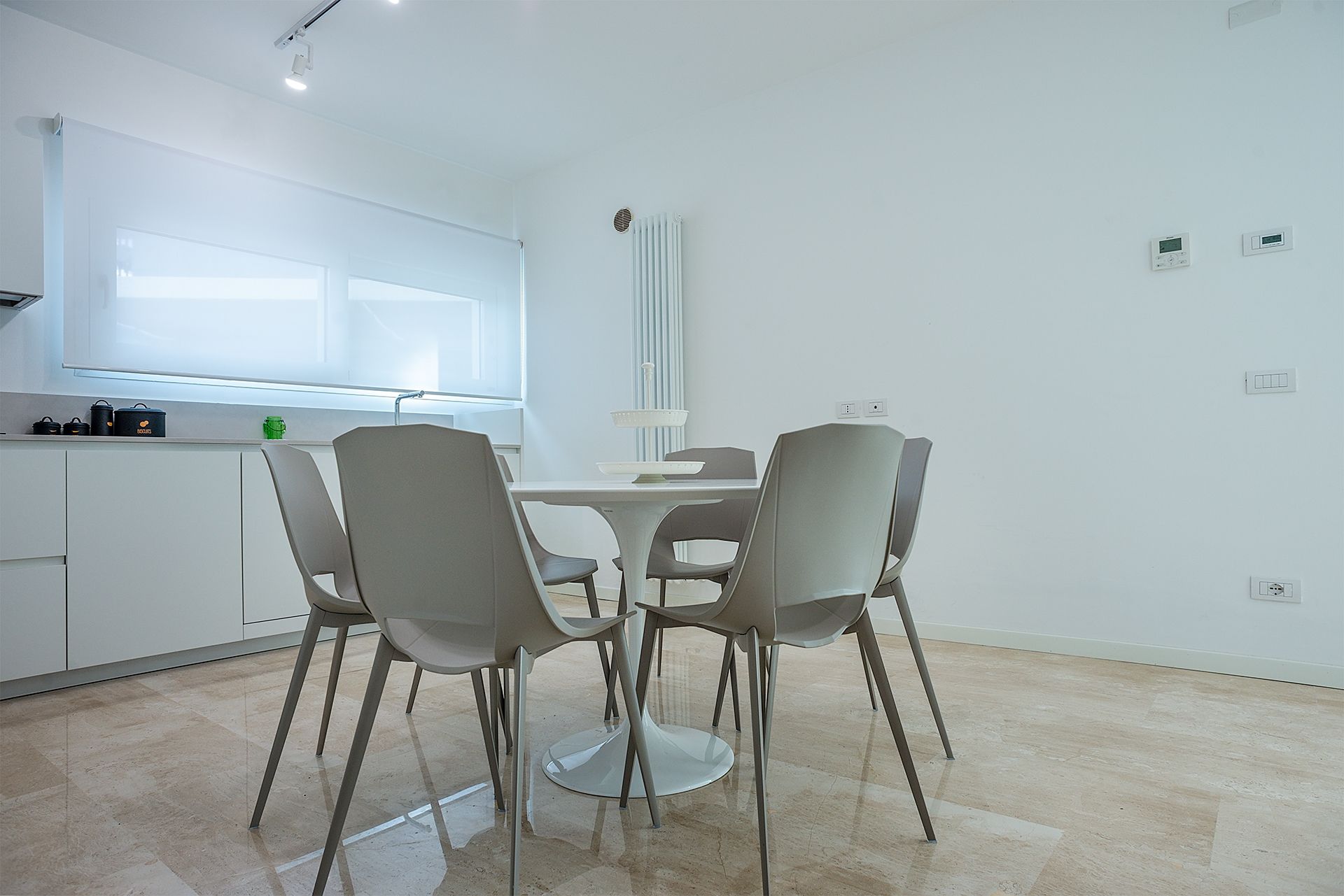 Uma cozinha moderna com armários brancos, uma mesa de jantar com quatro cadeiras e um radiador na parede.