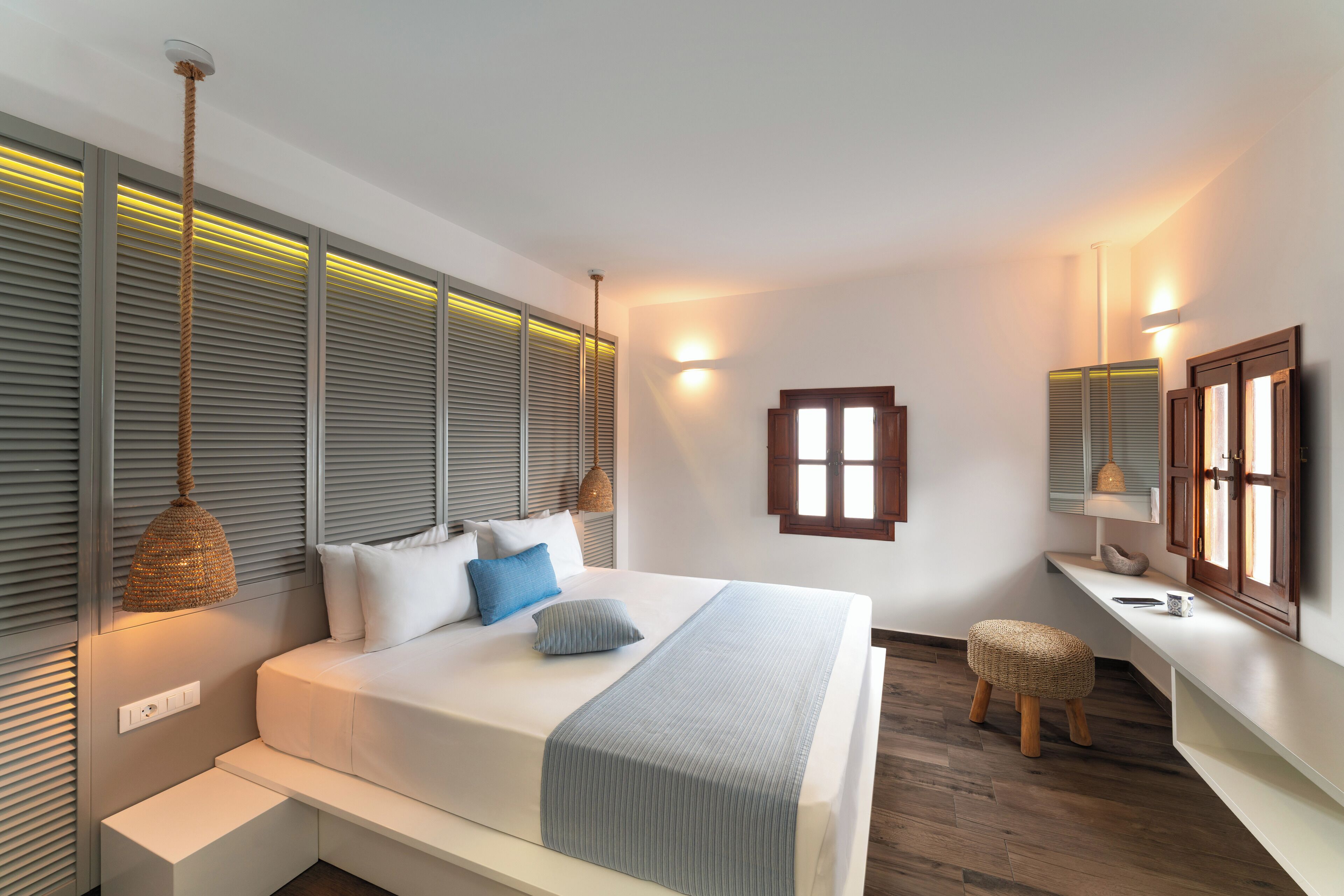 Quarto com cama grande, mesas de cabeceira, luminária pendente e janela com persianas.