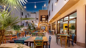 Uma área de refeições ao ar livre com várias mesas e cadeiras, rodeada por plantas e um edifício com grandes janelas.