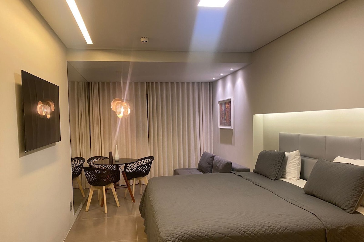 Quarto de hotel moderno com uma cama grande, área de refeições com cadeiras e vista para a cidade pela janela.