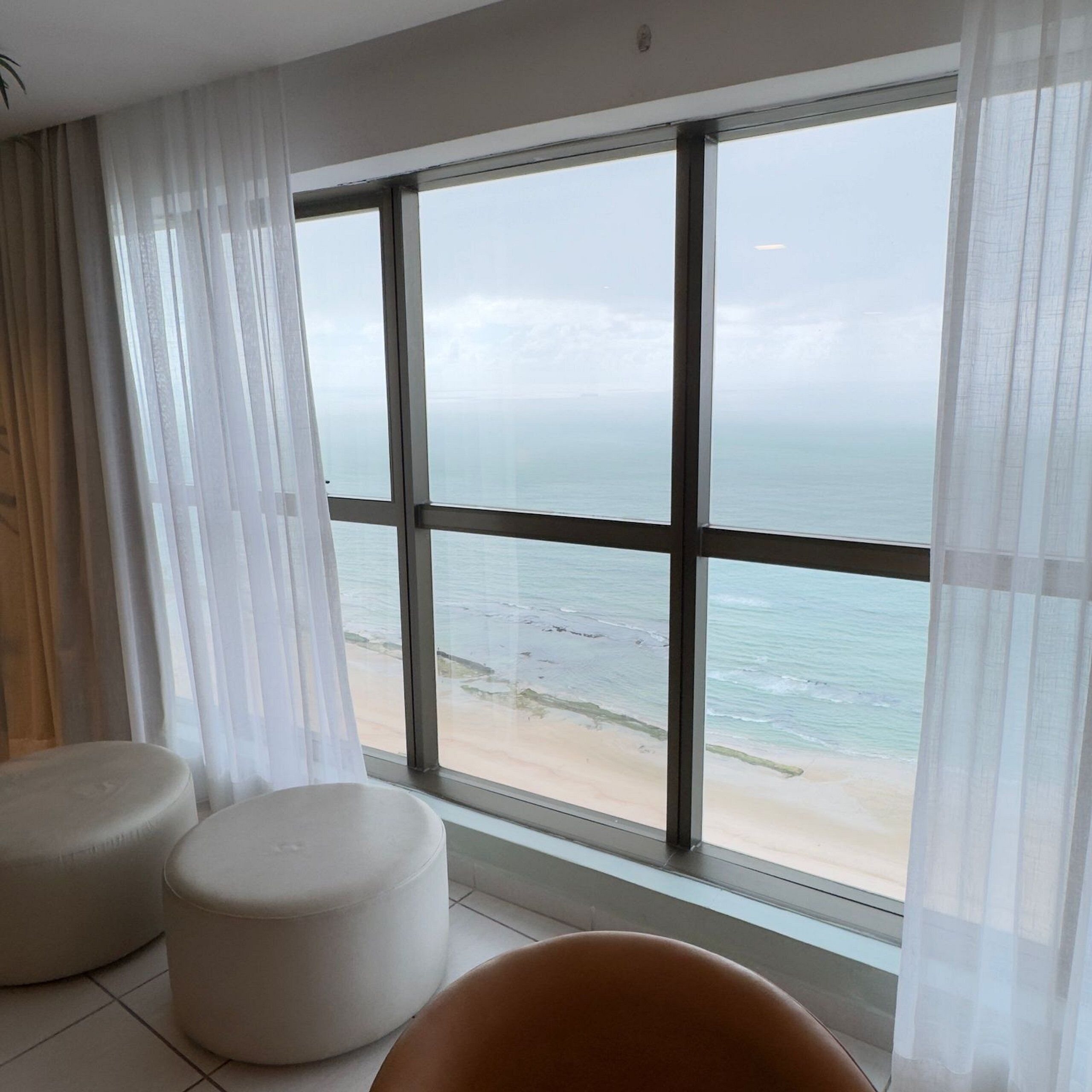 Quarto com uma grande janela de frente para a praia. Há dois pufes redondos e uma única cadeira.