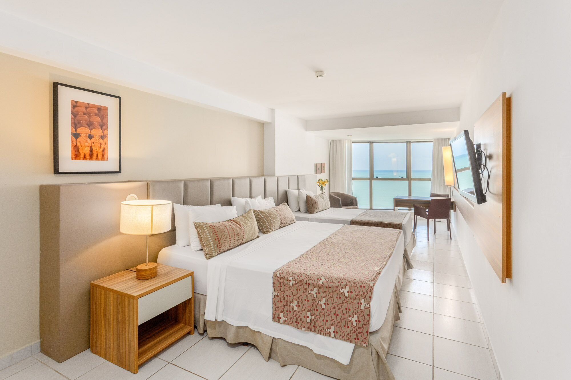 Quarto de hotel com duas camas, uma escrivaninha, uma cadeira, uma televisão e uma janela grande com vista para o mar.