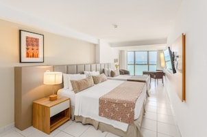 Quarto de hotel com duas camas, uma escrivaninha, uma cadeira, uma televisão e uma janela grande com vista para o mar.