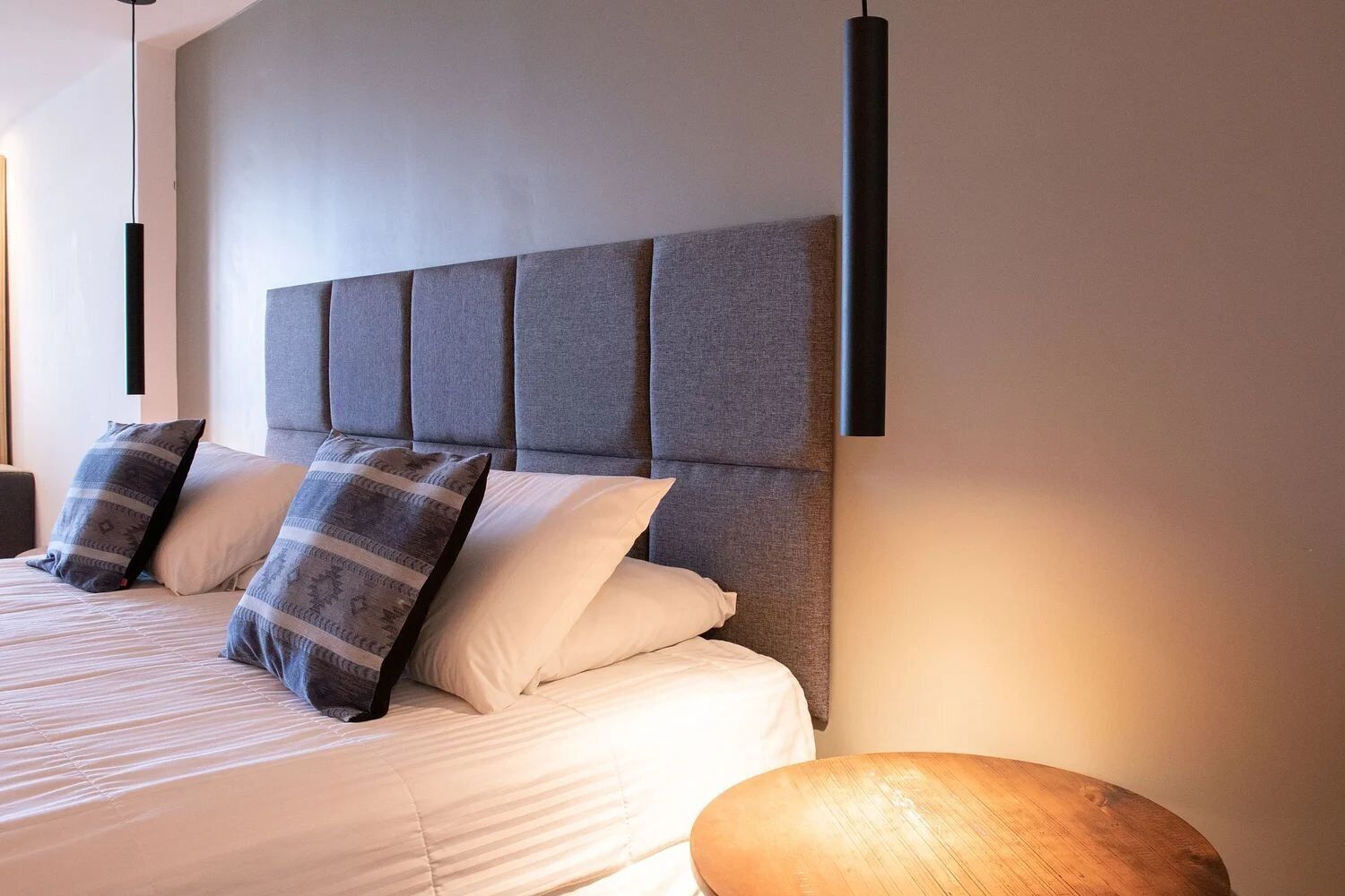 Quarto de hotel com uma cama grande, cabeceira com vários painéis e uma mesa de madeira redonda.