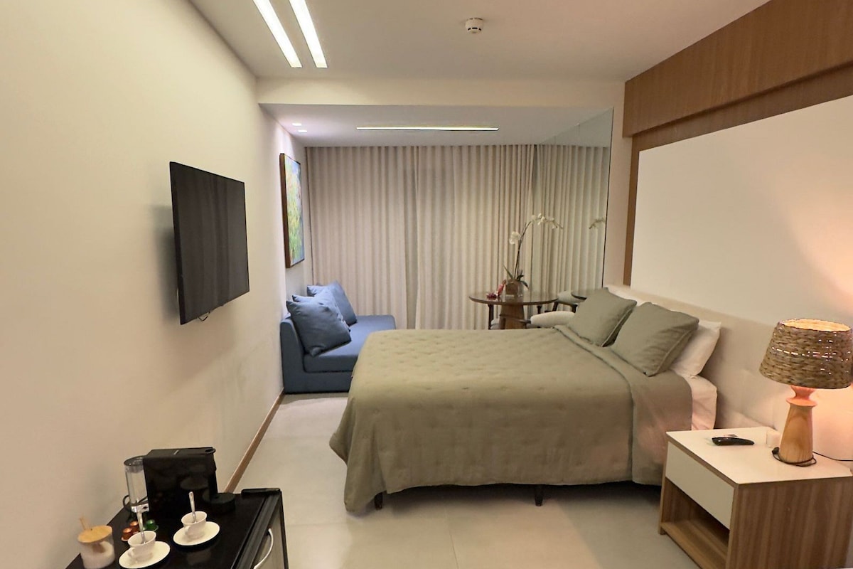 Quarto de hotel moderno com cama, sofá, escrivaninha com luminária e televisão na parede.