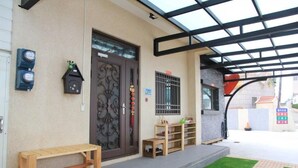 Entrada de um edifício com um banco de madeira, uma porta de metal com detalhes decorativos e um pequeno móvel com objetos.