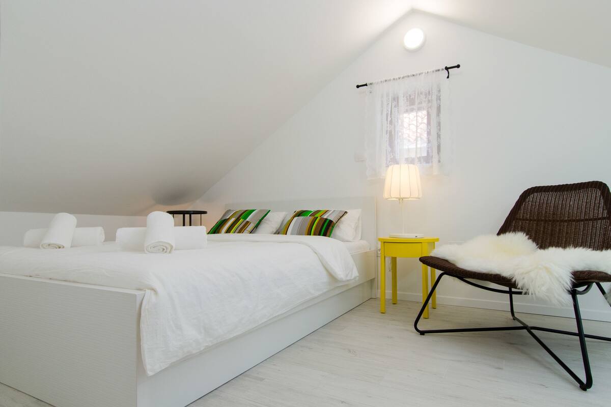 Quarto com uma cama, uma cadeira, uma mesinha e uma janela com cortina.
