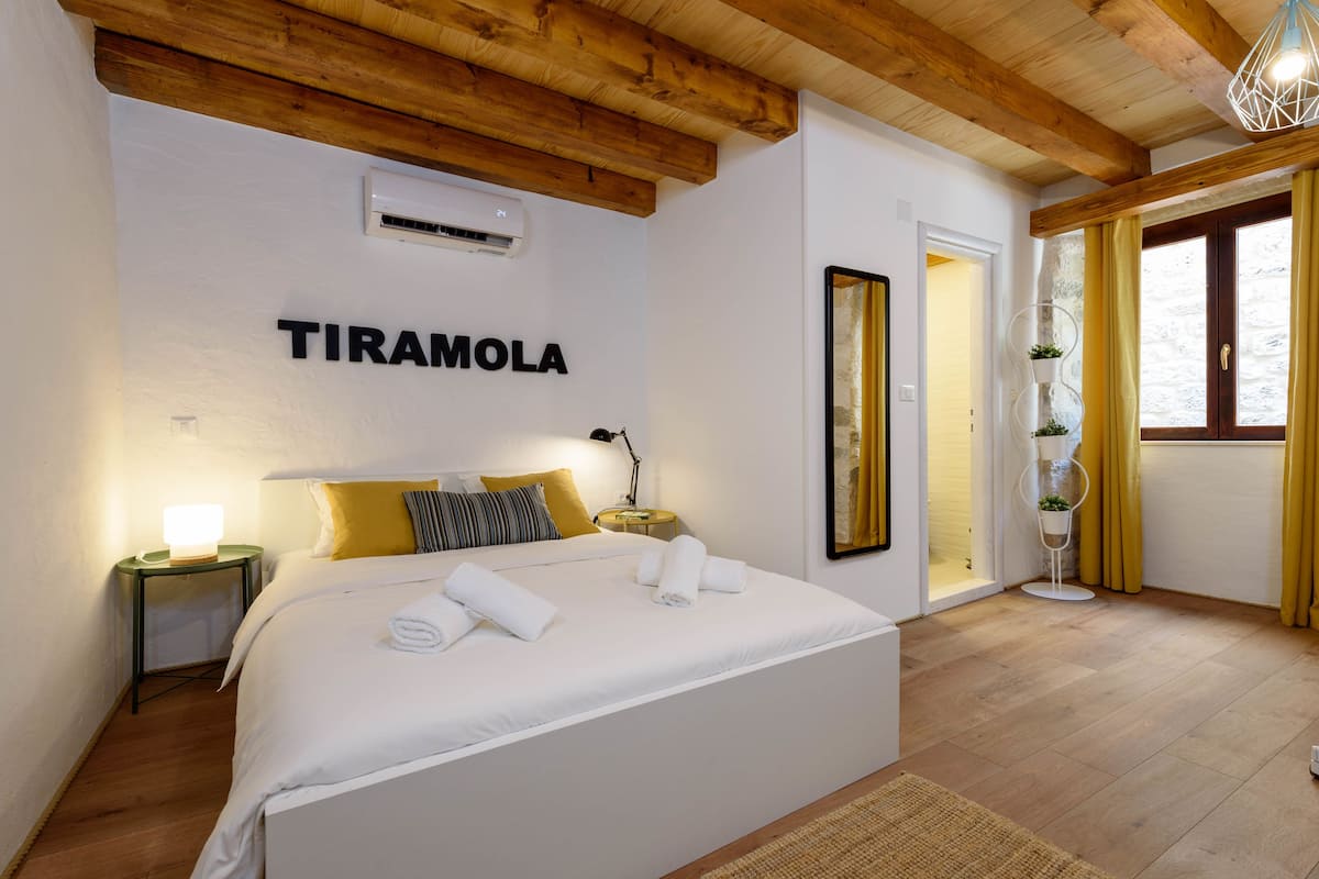 Quarto com cama, mesa de cabeceira, luminária, ar-condicionado fixado na parede e janela com cortinas.