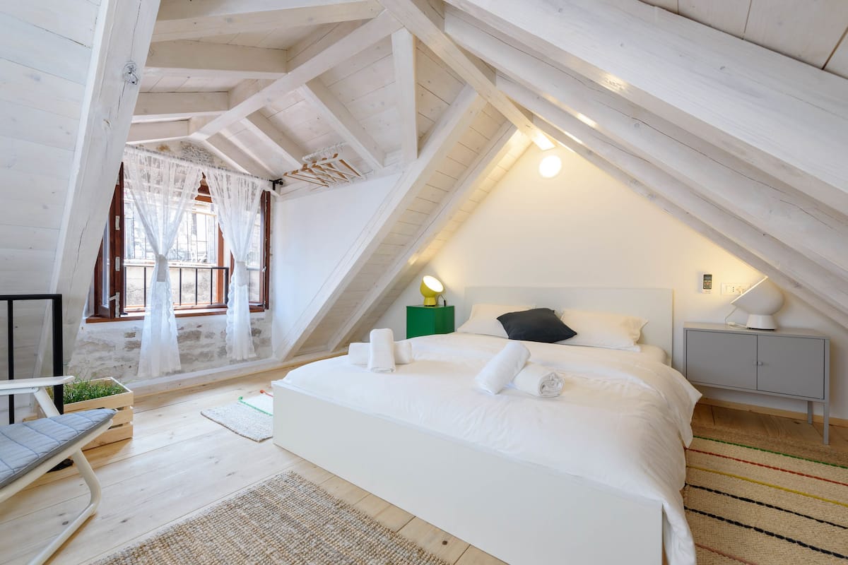 Quarto com teto inclinado, cama com roupa branca, mesa de cabeceira com abajur e vista para uma varanda com cortinas.