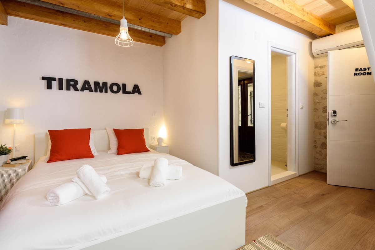 Quarto de hotel com uma cama, dois travesseiros vermelhos, roupa de cama branca, um espelho fixado na parede e uma placa com a inscrição "TIRAMOLA".