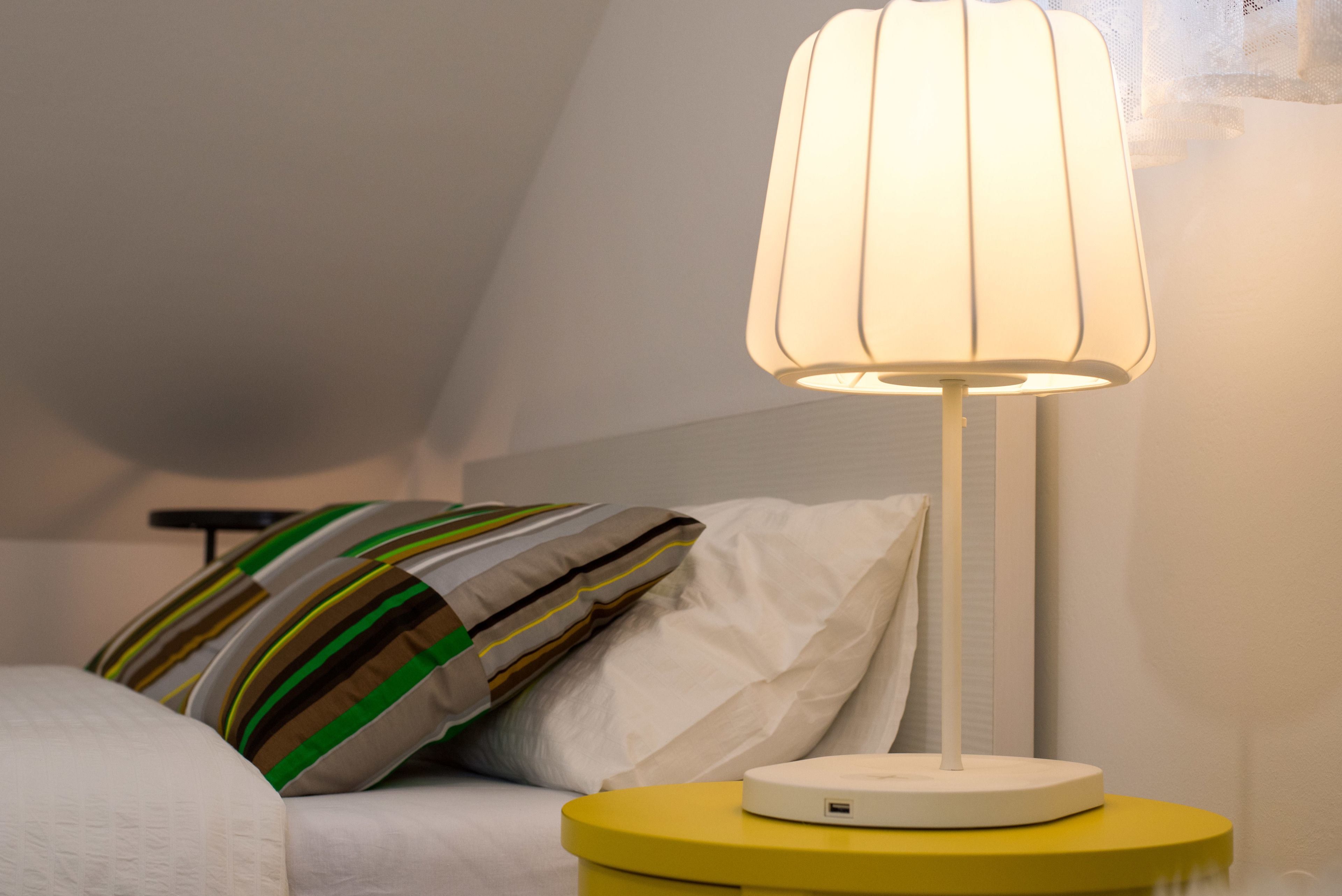 Uma cama com travesseiro branco e um travesseiro listrado, uma mesinha de cabeceira amarela com abajur branco, e uma parede branca ao fundo.