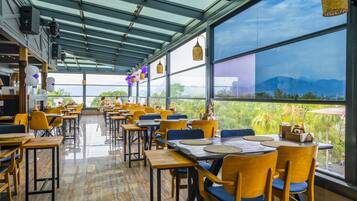 Um restaurante espaçoso com pé-direito alto e grandes janelas que oferecem vista para vegetação e montanhas. As mesas estão dispostas de forma organizada, com cadeiras amarelas.