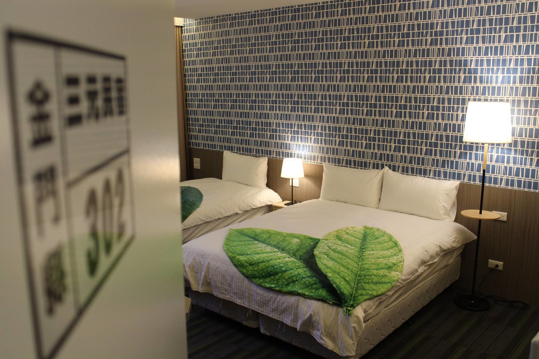 Quarto de hotel com duas camas, parede com padrão azul e branco e travesseiro em formato de folha verde.